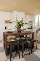 Uma cozinha com mesa de madeira, banquetas, um vaso com flores e diversos utensílios.
