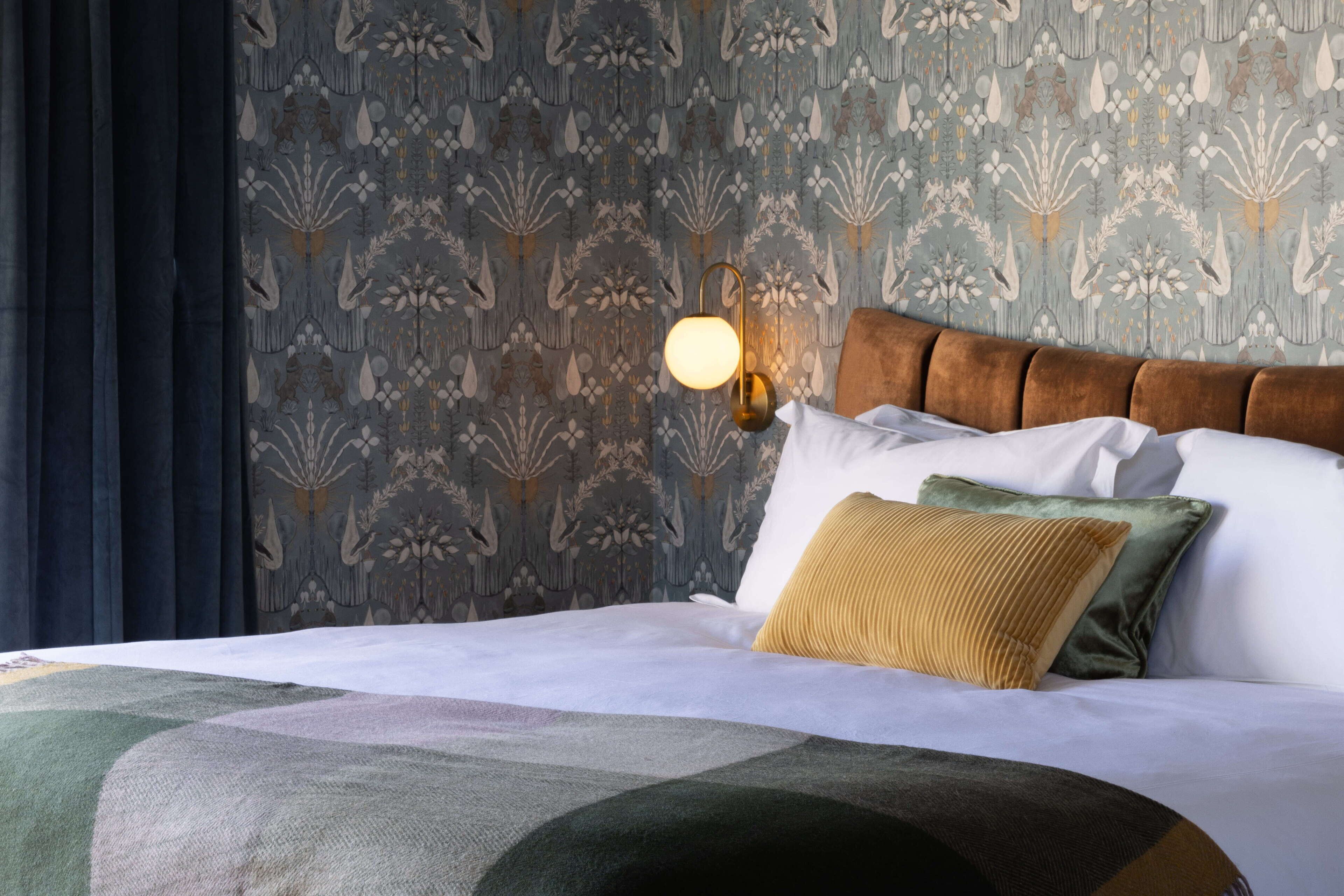 Cama bem arrumada com lençóis brancos e várias almofadas, papel de parede estampado e luminária fixada na parede.