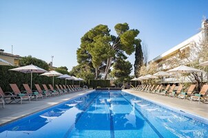 Piscina com espreguiçadeiras e guarda-sóis, cercada por árvores e edifícios.