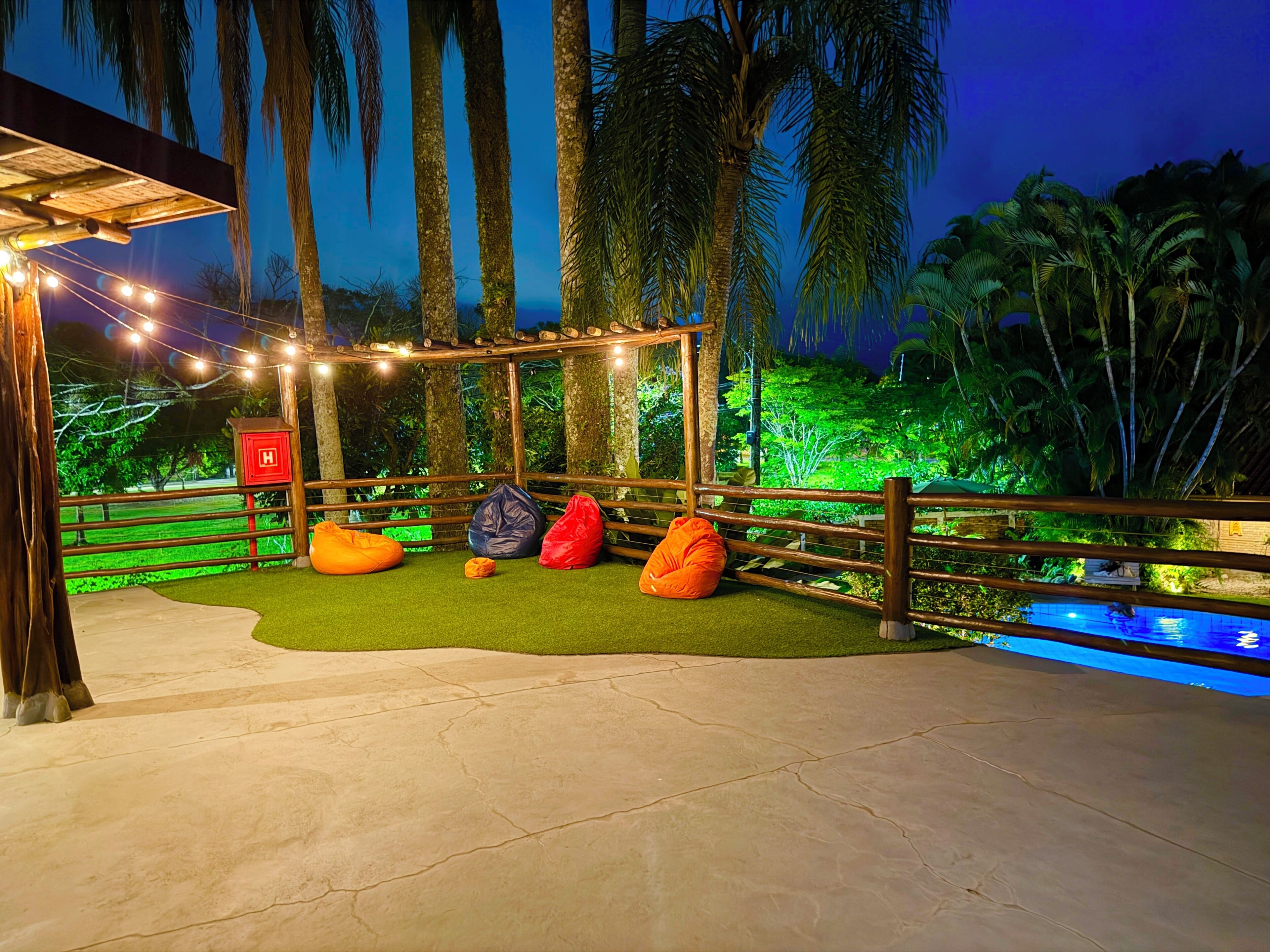 Uma área de estar ao ar livre à noite, com luzes de corda, piscina e palmeiras.