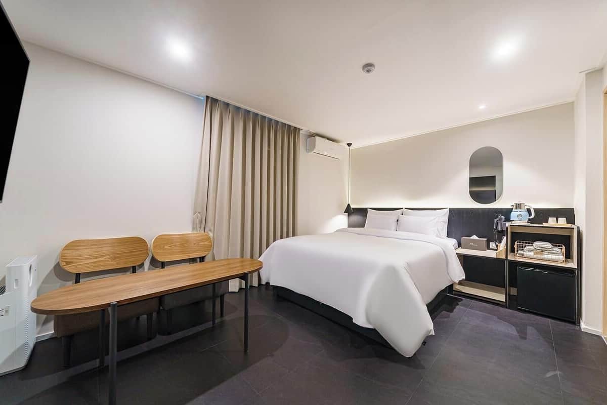 Quarto de hotel moderno com cama, mesa de cabeceira, luminária e um pequeno espaço de estar com um banco.