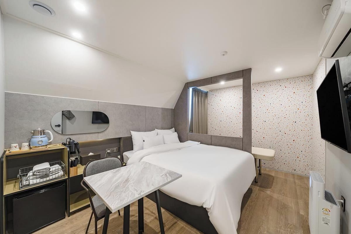 Quarto de hotel moderno com cama, escrivaninha, cadeira, mesa pequena, televisão e mini-geladeira.