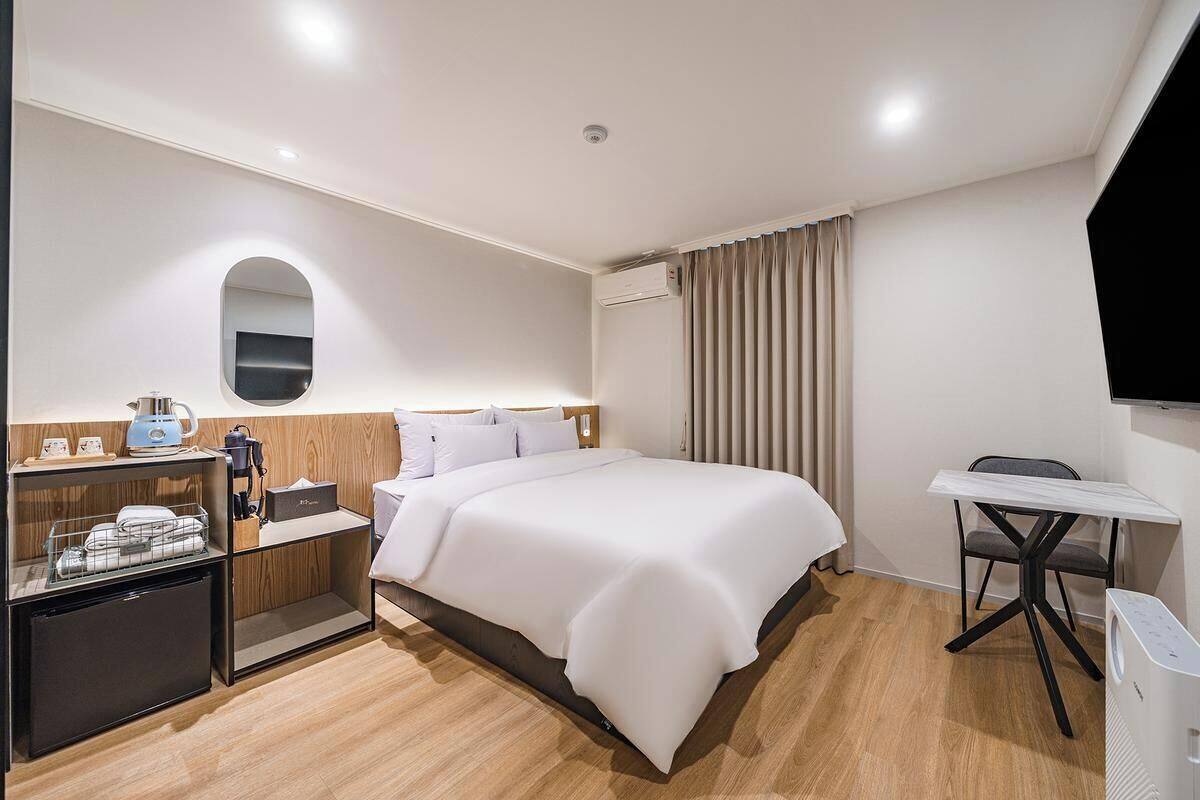Quarto de hotel com cama, escrivaninha, cadeira, pequeno frigorífico, máquina de café e televisão fixada na parede.