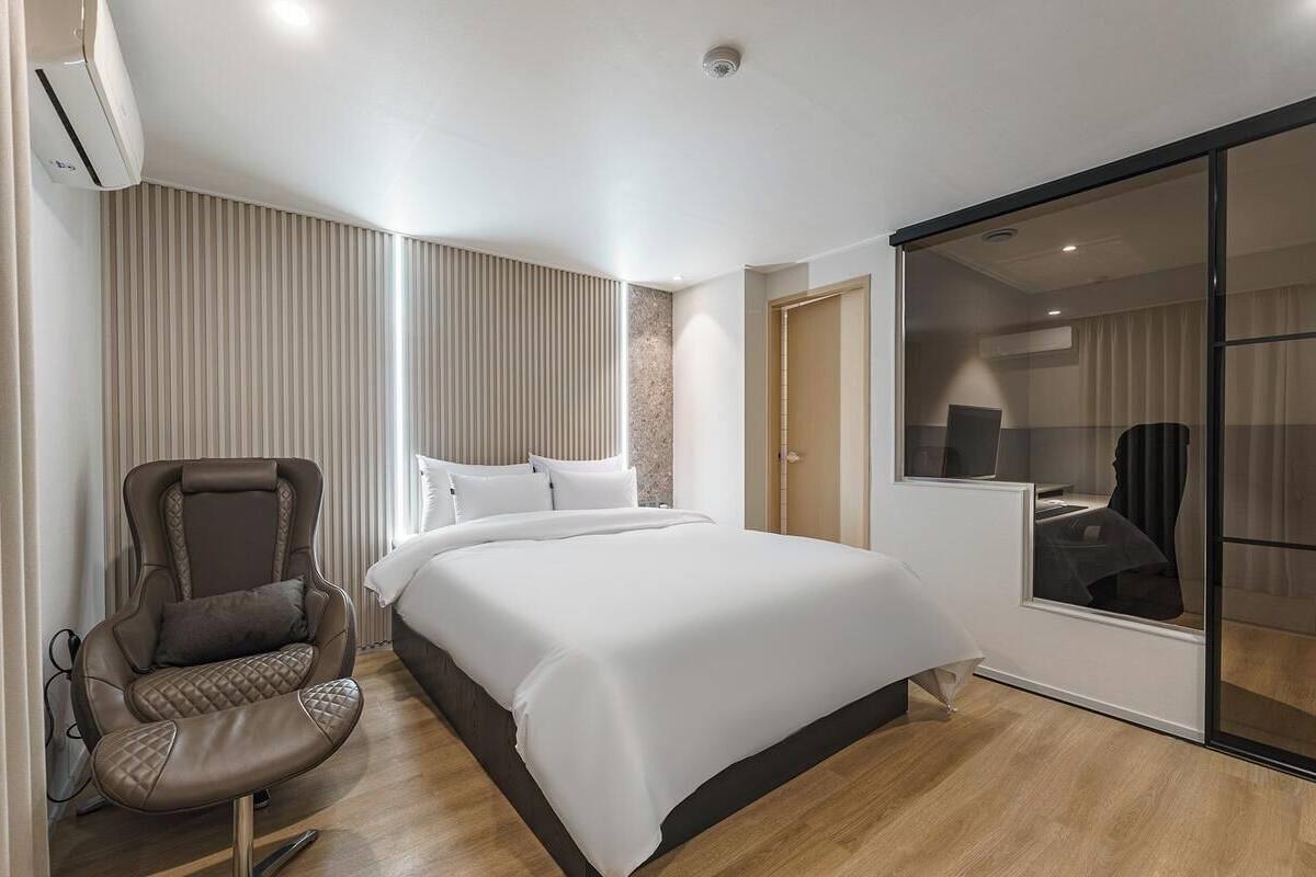 Quarto de hotel moderno com uma cama grande, uma poltrona reclinável, uma porta de vidro deslizante para uma varanda e um ar condicionado fixado na parede.