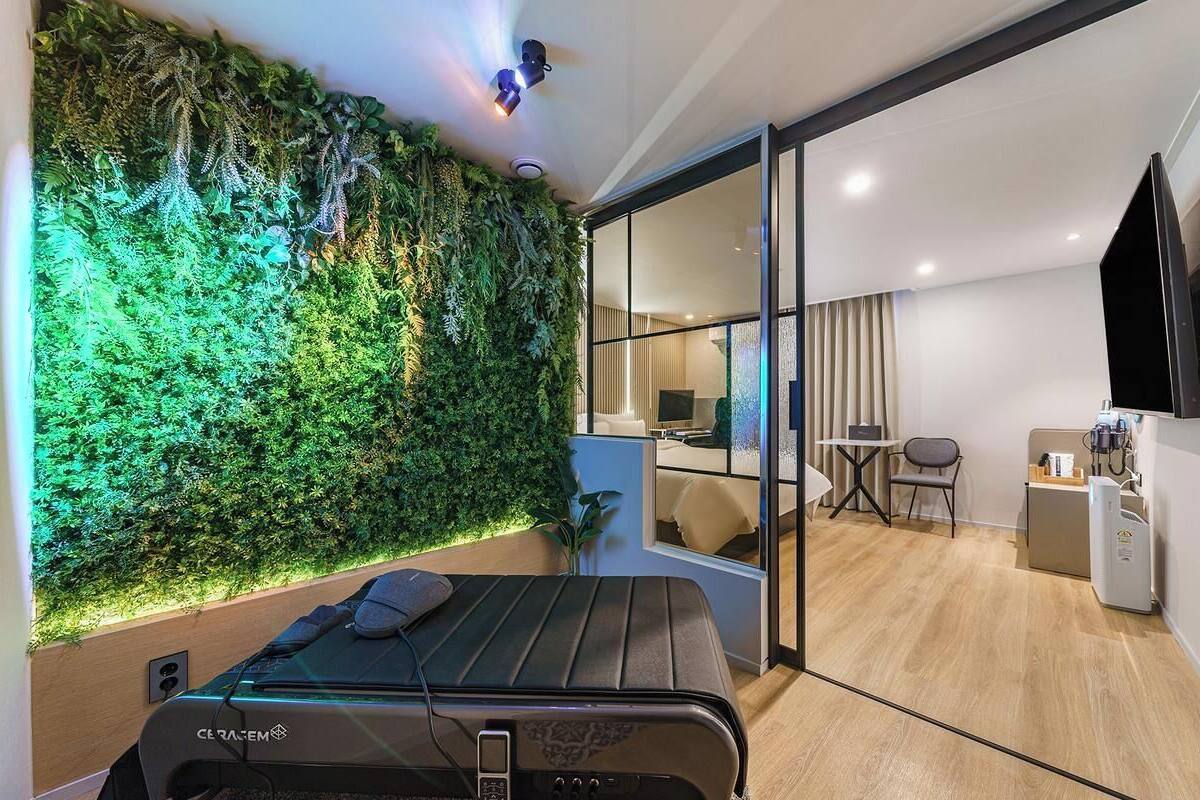 Quarto de hotel moderno com parede verde, mesa de massagem, TV e cozinha compacta.