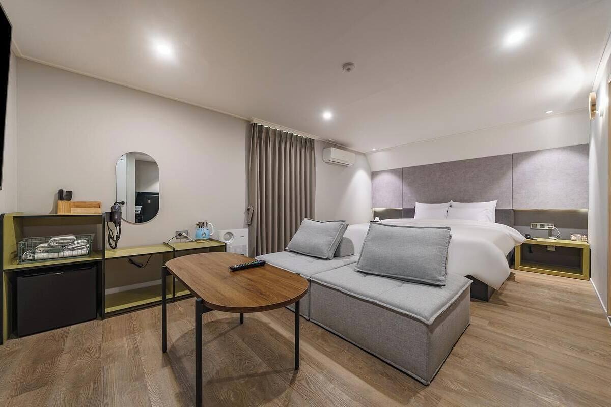 Quarto de hotel moderno com sofá-cama, pequena escrivaninha com cadeira, mesa de cabeceira e televisão na parede.