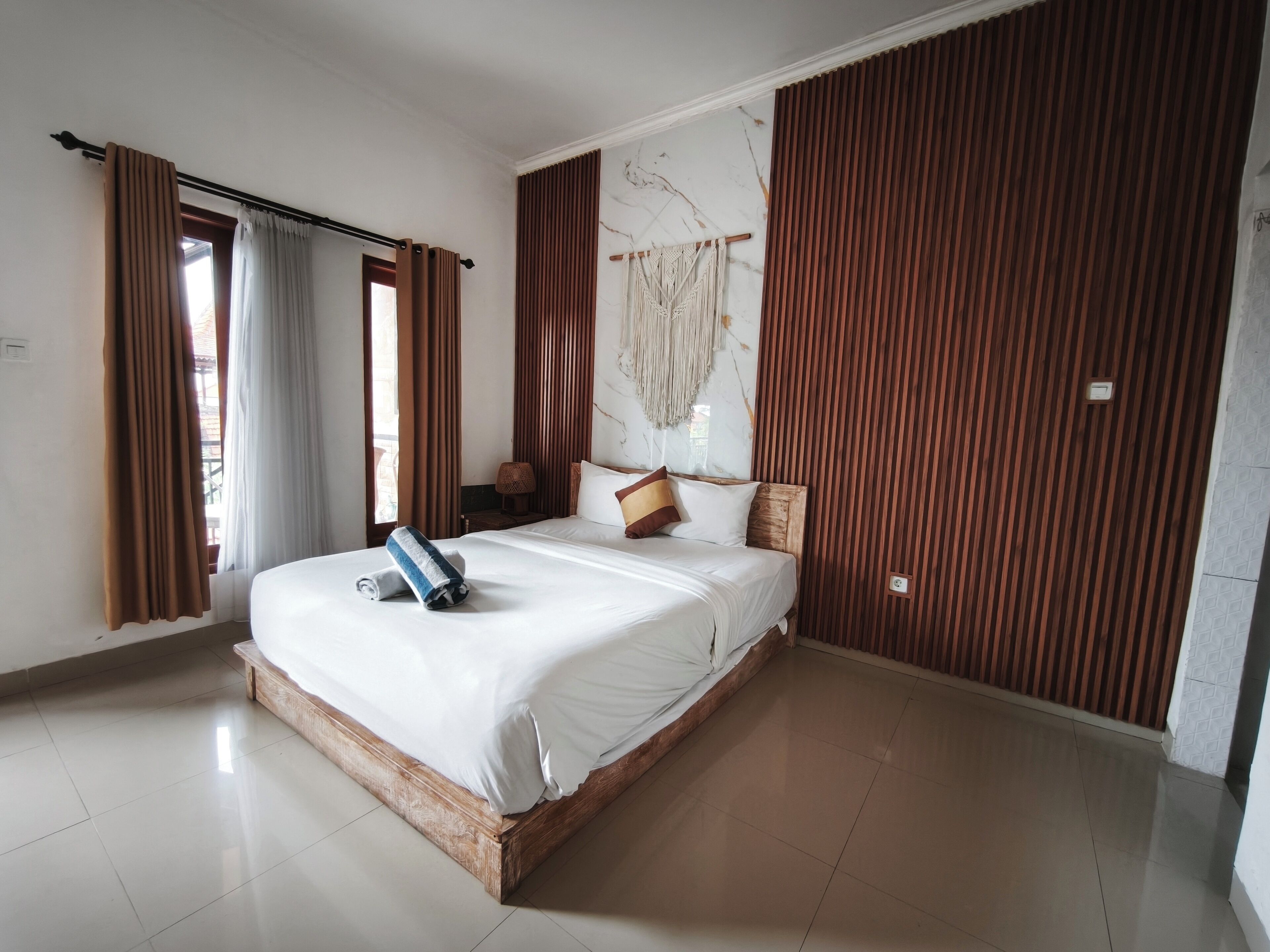Quarto de hotel com uma cama grande, cabeceira de madeira, uma peça de macramé na parede e vista do exterior através de uma janela com cortinas.