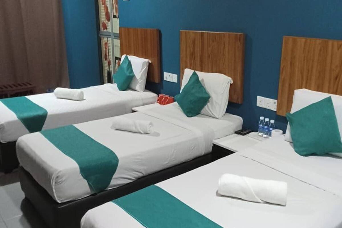 Quarto de hotel com três camas de solteiro, cada uma com colcha branca e verde, cabeceira de madeira e ar condicionado fixado na parede.