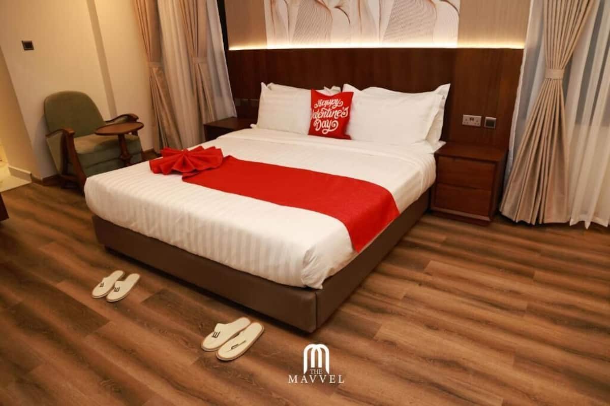 Quarto de hotel com uma cama grande, um cobertor vermelho, chinelos no chão e um travesseiro com a mensagem 'Feliz Dia dos Namorados'.