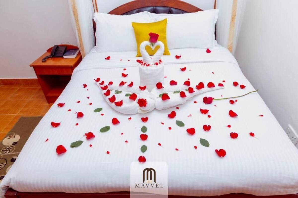Cama bem arrumada com pétalas de rosa e um arranjo de toalha em formato de coração, um telefone na mesinha de cabeceira e um travesseiro amarelo sobre um cobreleite branco.