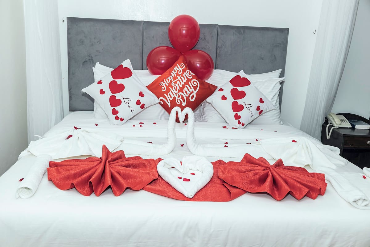 Uma cama bem arrumada com arranjo de toalhas em formato de coração, almofadas vermelhas e brancas e um balão decorativo com a mensagem 'Happy Valentine's Day'.