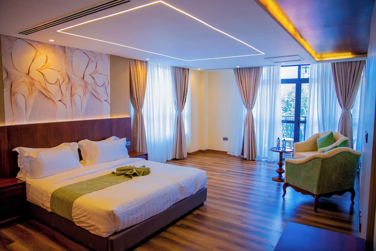 Quarto de hotel com uma cama grande, uma poltrona verde e vista para o exterior pelas janelas.