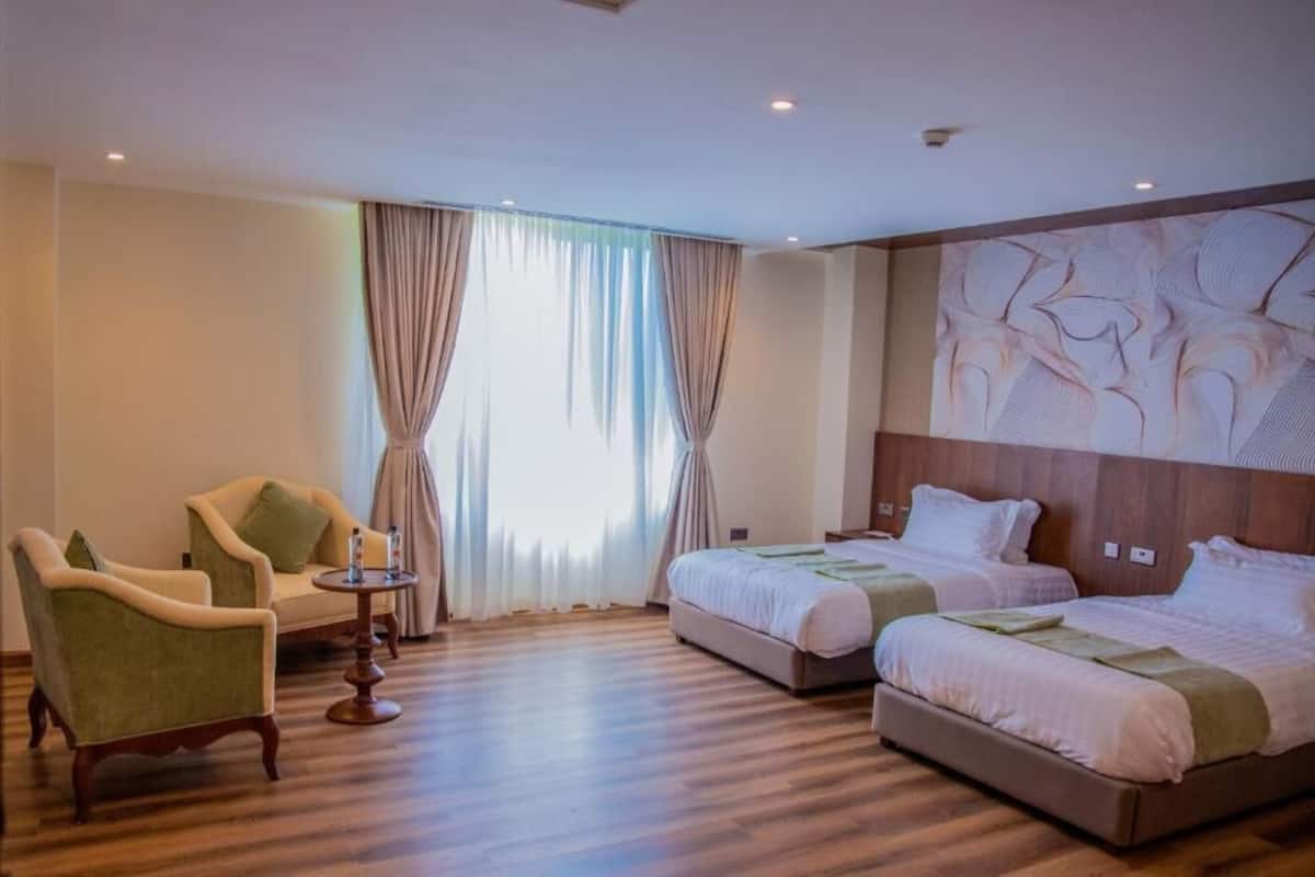Quarto de hotel com duas camas, área de estar com uma cadeira e uma mesinha, e uma grande obra de arte abstrata na parede.