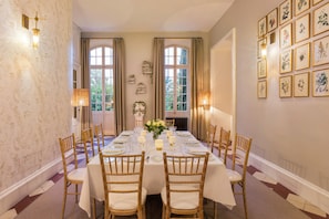 Uma sala de jantar formal com uma mesa longa posta para seis pessoas, com janelas altas com cortinas e impressões botânicas emolduradas nas paredes.