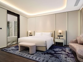 Quarto de hotel moderno com uma cama grande, mesas de cabeceira, um banco e um tapete estampado.