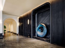 Um hall de hotel moderno com piso de mármore, painéis de madeira escura e uma grande escultura abstrata.