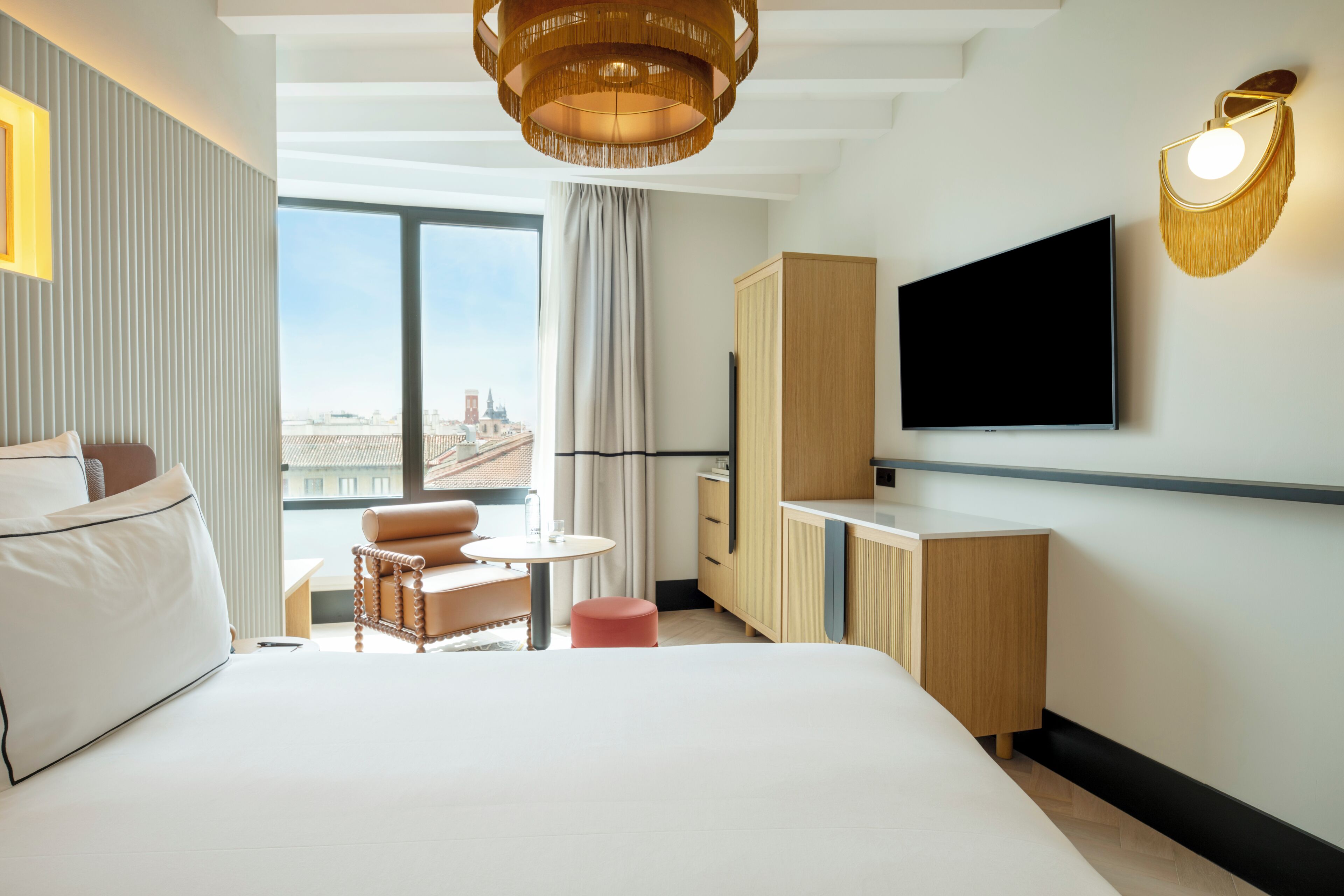 Quarto de hotel moderno com uma cama grande, televisão na parede, um pequeno espaço de refeições e vista para a cidade.
