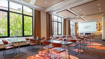 Sala de conferências com mesas, cadeiras, tela de projetor e grandes janelas com vista para edifícios e árvores.