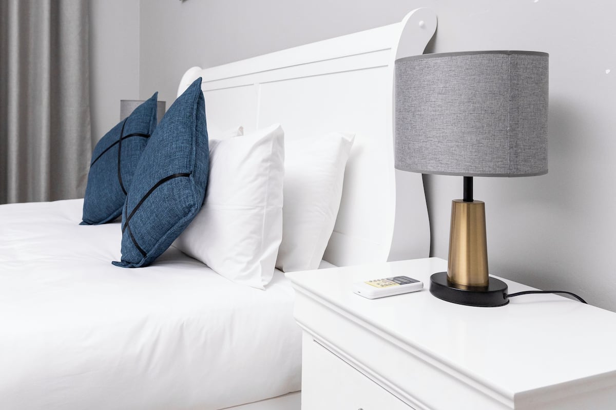 Cama bem arrumada com roupa de cama branca e almofadas azuis, mesa de cabeceira com abajur e controle remoto sobre a mesa.