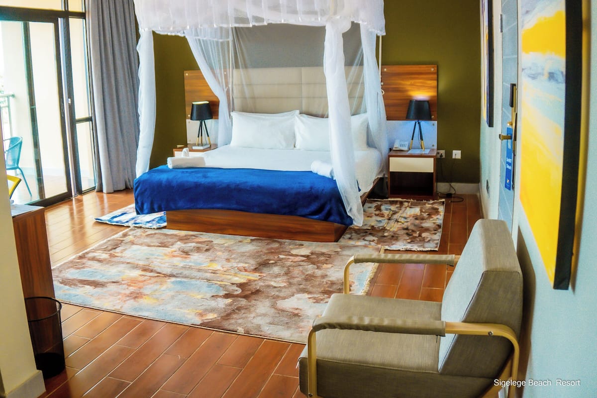 Quarto com cama de dossel, uma cadeira, mesa de cabeceira com abajur e vista para a praia.