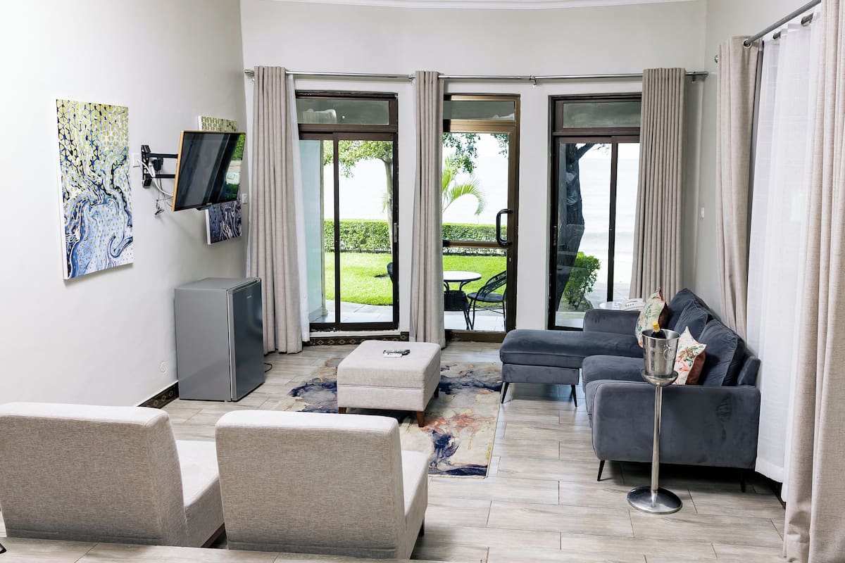 Uma sala de estar moderna com um sofá cinza, uma televisão pequena e uma mesa de centro. Duas portas de vidro corremoras dão acesso a uma área externa com vista para o verde.