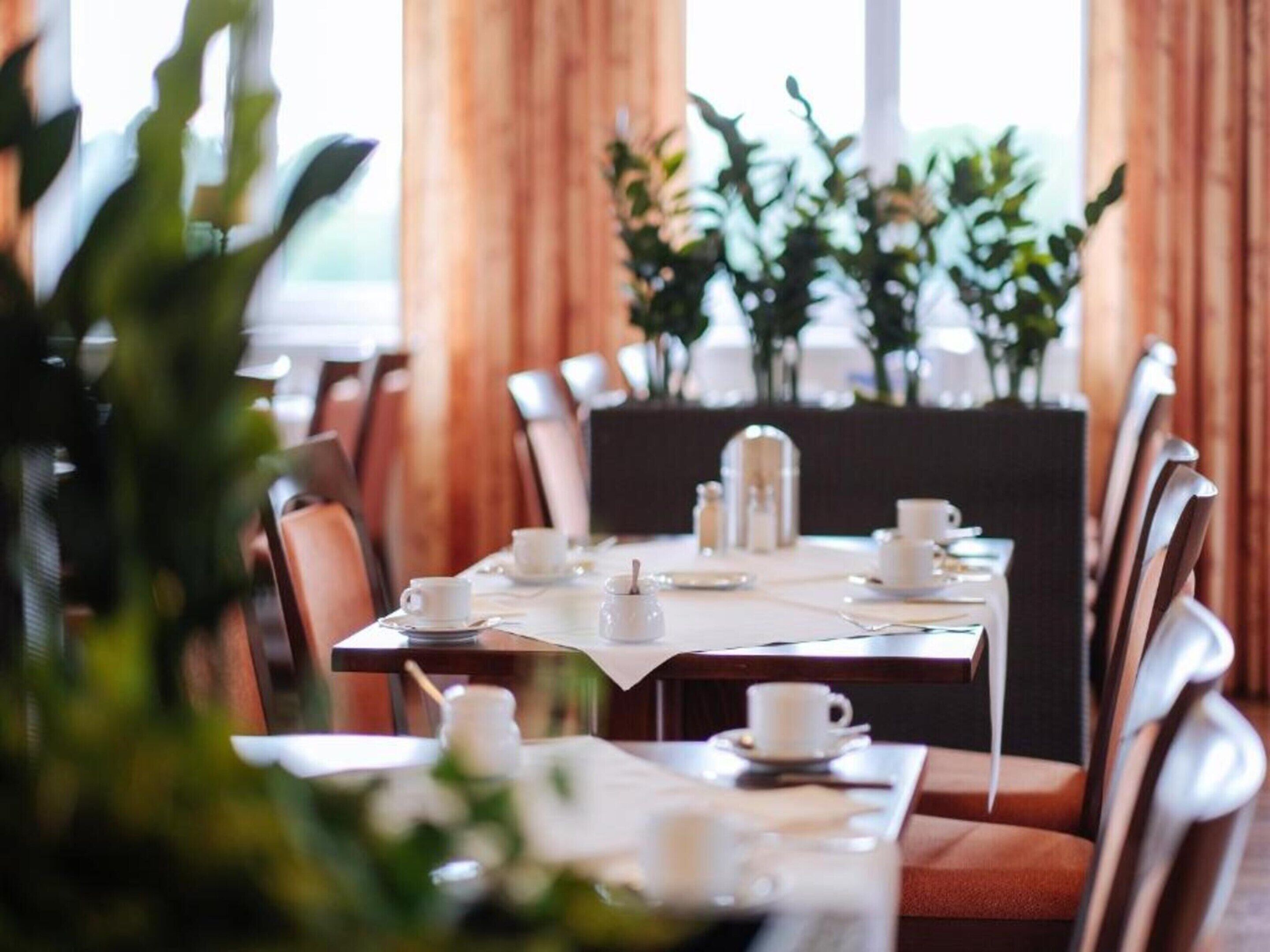 Uma sala de jantar com mesas preparadas para o serviço, cadeiras e uma grande janela com cortinas.