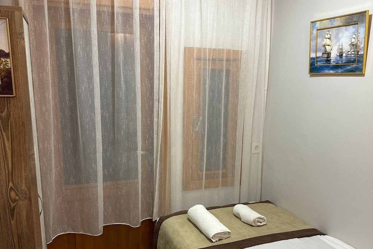 Quarto de hotel com cama, uma cadeira de madeira, janela com cortinas leves e um quadro na parede.