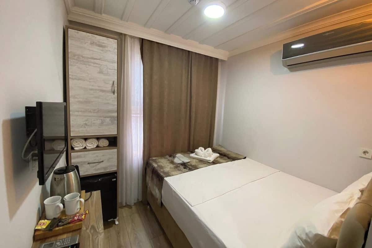 Quarto de hotel com cama, mesa de cabeceira com chaleira, chávenas e comando. Existe um guarda-roupa com espelho e cortina.