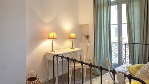 Quarto com cama, escrivaninha com duas luminárias e janela com cortinas.