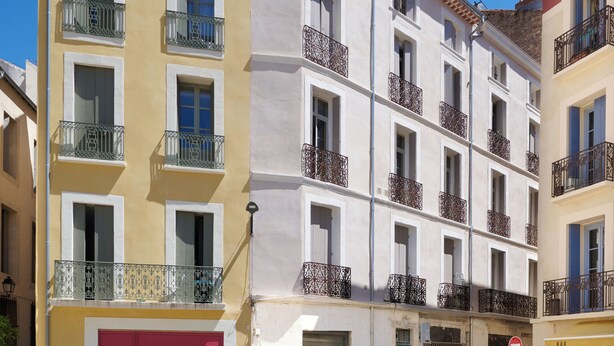 Um edifício com várias andares, varandas, uma porta vermelha e um pátio de paralelepípedos.