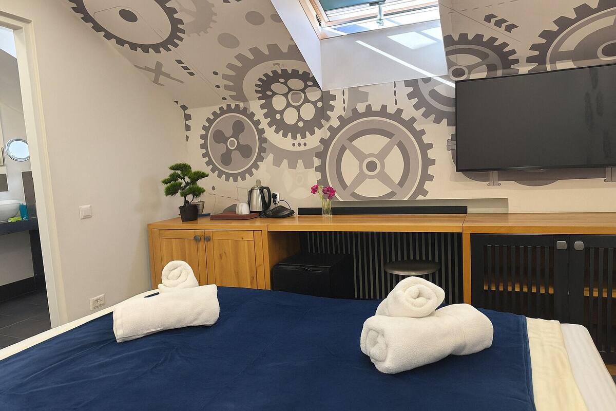 Quarto de hotel com cama, duas toalhas, escrivaninha com uma planta, chaleira e televisão.