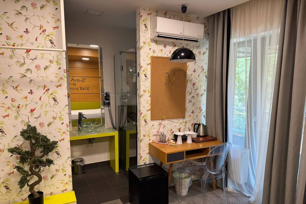Quarto de hotel pequeno e funcional, com cozinha compacta, escrivaninha, cadeira, planta em vaso e uma janela grande com cortinas.