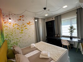 Quarto de hotel com cama, escrivaninha, cadeira, planta em vaso e janela com cortinas.