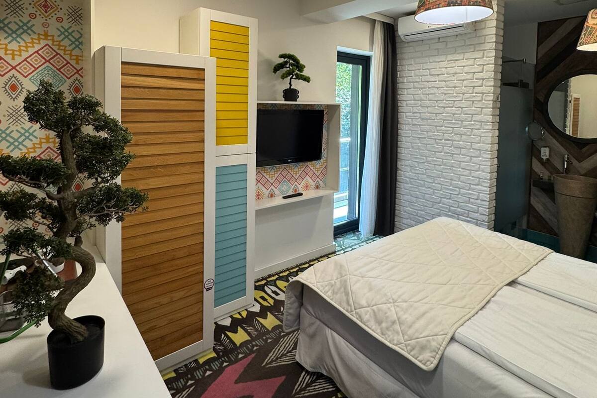 Quarto moderno com cama, televisão, planta em vaso e tapete estampado.