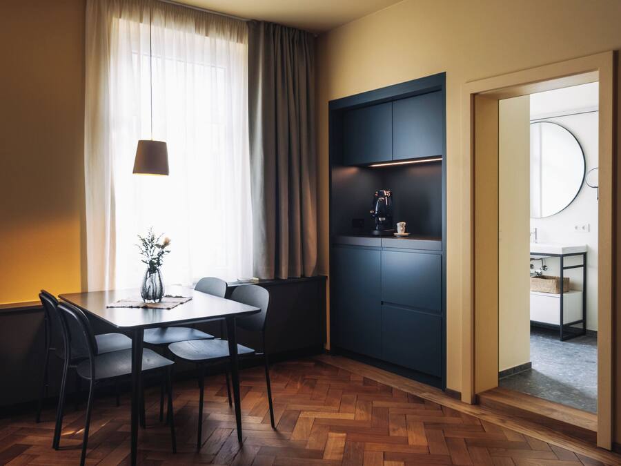 Uma área de jantar moderna com piso de madeira em padrão herringbone, uma mesa com cadeiras, um vaso com flores e um armário azul escuro com uma máquina de café.
