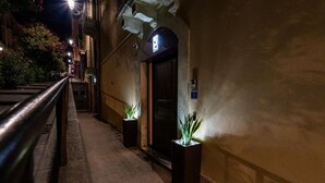 Vista noturna da fachada de um edifício, com guarda-corrimão de metal, uma porta com uma placa acima e duas plantas em vasos na calçada.