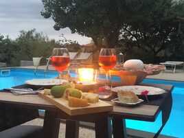 Mesa junto à piscina com um prato de frutas, duas taças de vinho tinto e um cesto de pão e queijo.