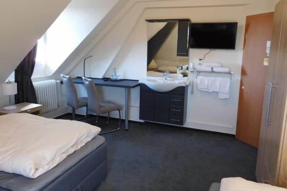 Quarto de hotel com cama, escrivaninha, cadeira, luminária de mesa, pia, espelho e televisão.
