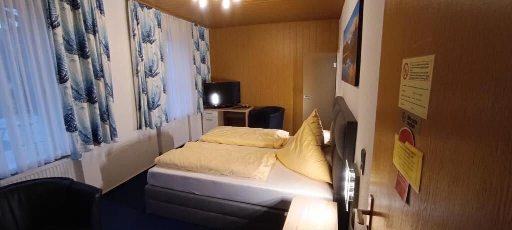 Quarto compacto com cama, escrivaninha, cadeira e televisão.