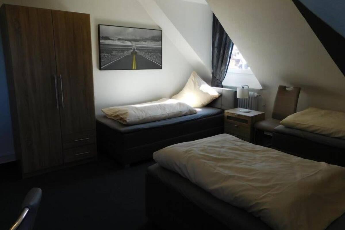 Um quarto pequeno e pouco iluminado, com uma cama de solteiro, uma mesinha de cabeceira, um guarda-roupa e um quadro na parede.