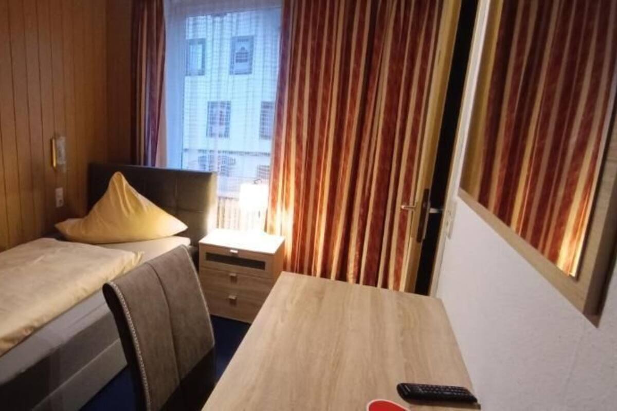 Quarto pequeno e compacto com uma cama de solteiro, mesa de cabeceira e janela com cortinas.