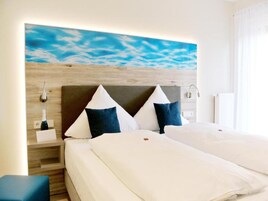 Quarto de hotel com cabeceira de madeira, cama com roupa de cama branca e uma obra de arte na parede retratando um céu azul.