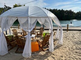 Uma cabana de praia com toldo branco, cadeiras de madeira e uma pequena mesa laranja.