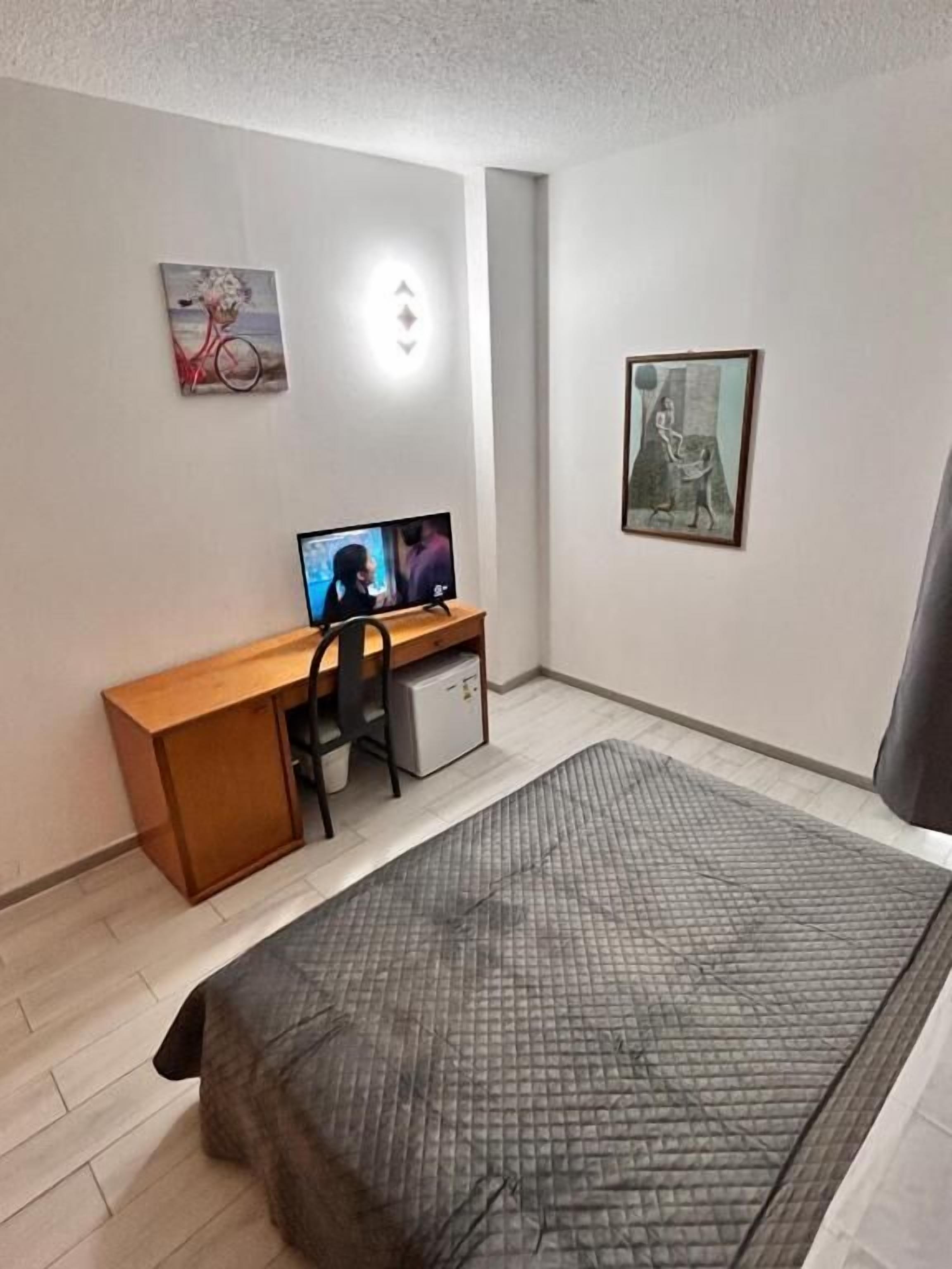 Quarto com cama, escrivaninha com computador e obras de arte nas paredes.