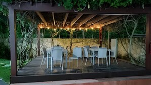 Um pátio ao ar livre com cadeiras e mesas brancas, sob uma pérgola com luzes de cordão.