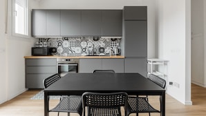 Uma cozinha moderna com armários cinza, bancada preta e painel decorativo. Há uma mesa de jantar com quatro cadeiras e um micro-ondas no balcão.