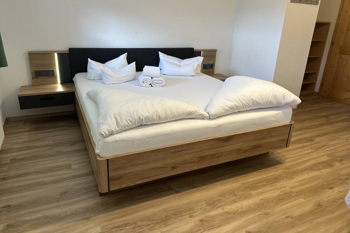 Cama bem arrumada com lençóis e almofadas brancas, cabeceira de madeira e mesa de cabeceira com abajur.