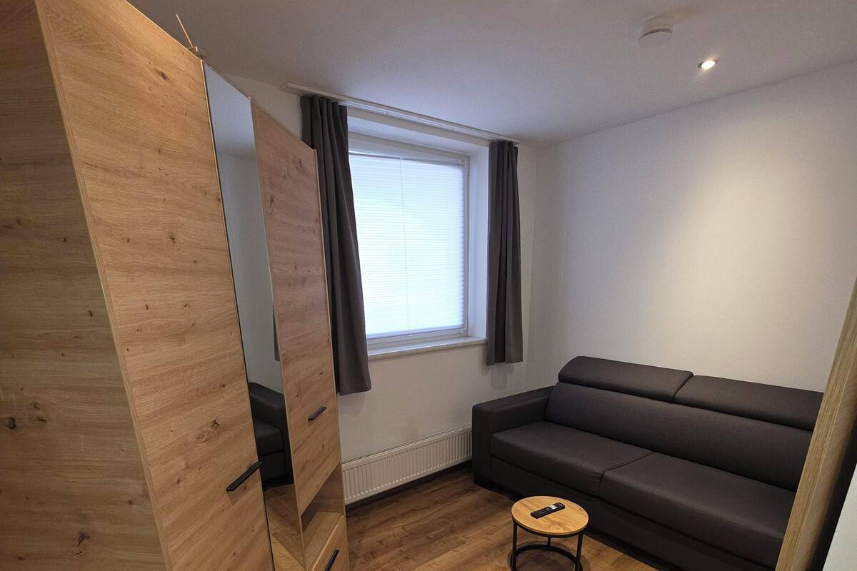 Quarto moderno com guarda-roupa de madeira, sofá cinza, mesa redonda pequena e janela com cortinas.