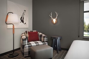 Um quarto com uma poltrona xadrez, um travesseiro vermelho, uma luminária, um chifre de veado fixado na parede, uma pequena mesa redonda e uma cama.