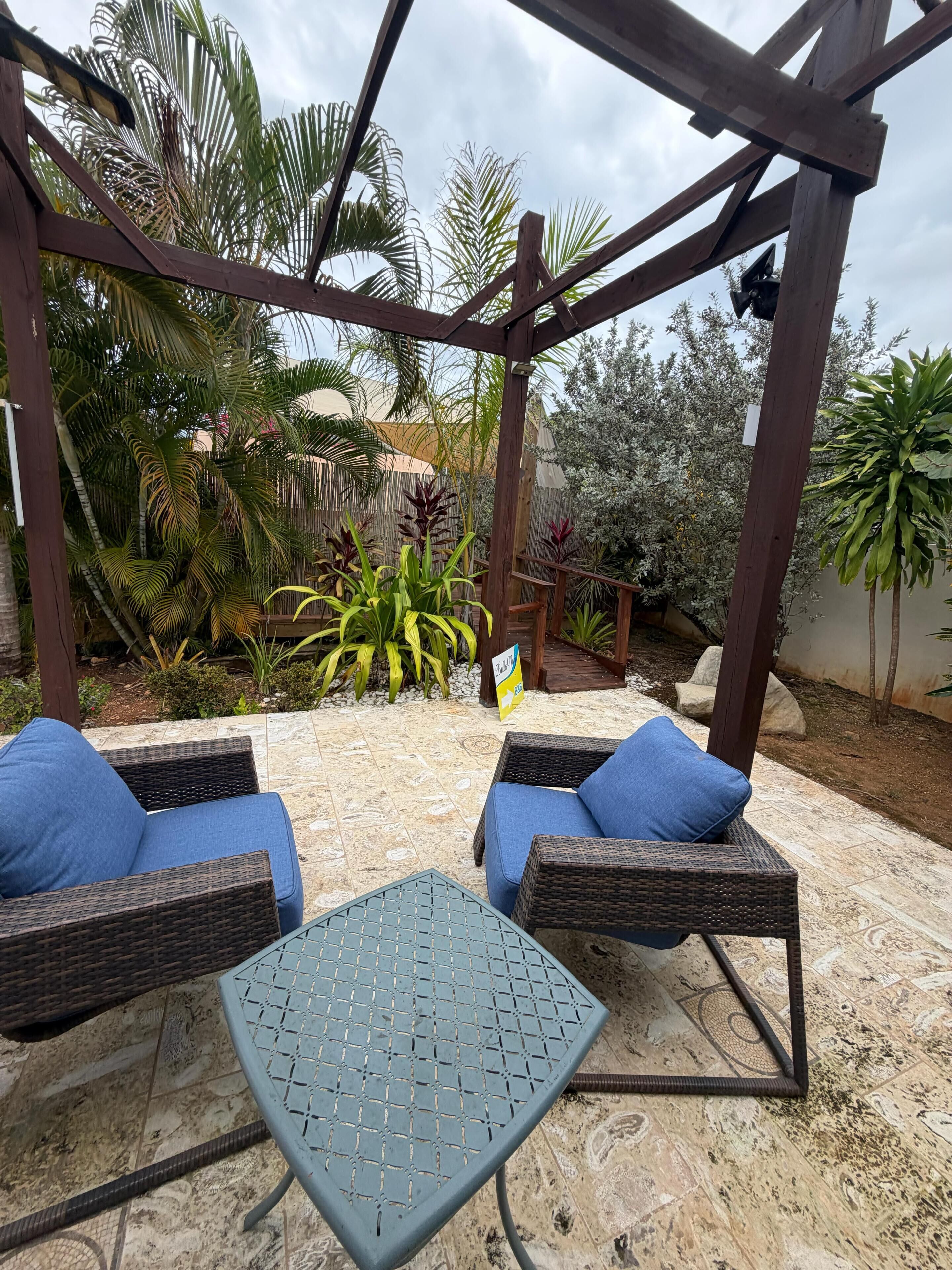 Pátio externo com mobiliário de vime e uma mesa de metal, rodeado por diversas plantas e uma estrutura de pérgula.